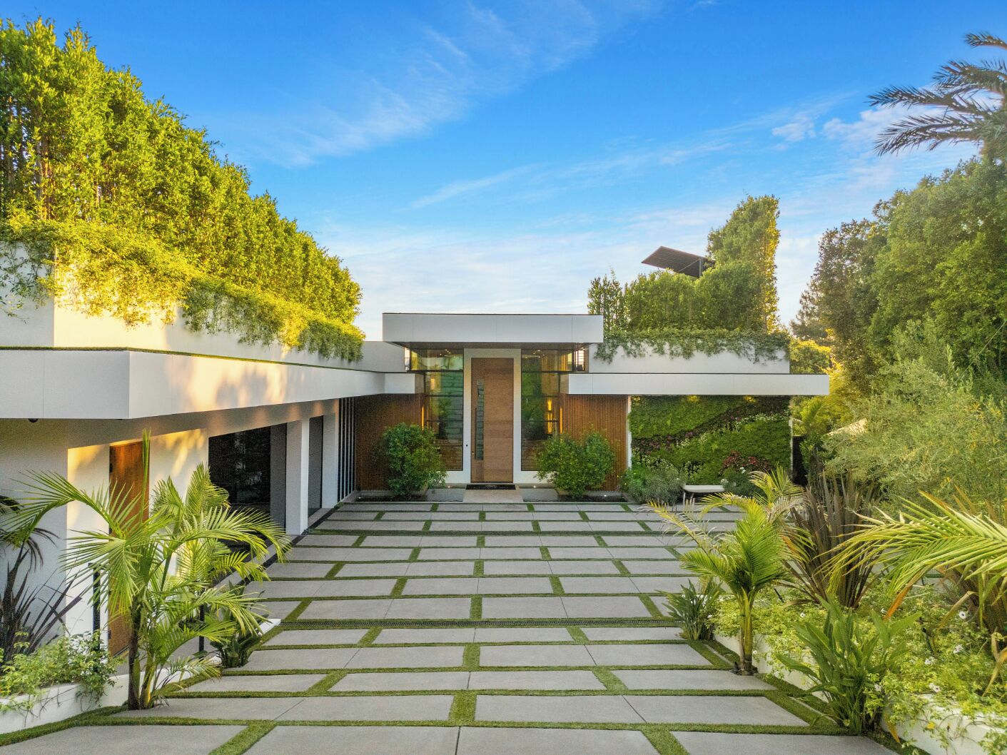 Uma casa moderna com entrada central, cercada por um jardim com diversas plantas e um caminho pavimentado.