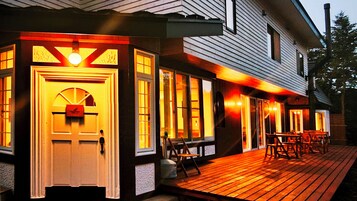 Deck de madeira com área de estar externa e porta de entrada iluminada.