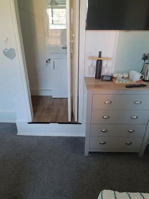 Quarto com piso de madeira, porta branca, penteadeira cinza com gavetas e uma pequena estante com uma chaleira e uma xícara.