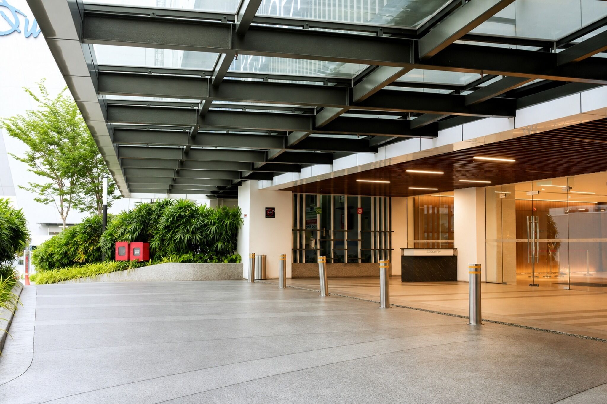 Entrada de um edifício moderno com toldo de vidro, balcão de segurança e área ajardinada com plantas.