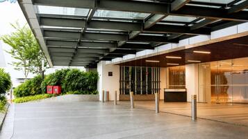 Entrada de um edifício moderno com toldo de vidro, balcão de segurança e área ajardinada com plantas.