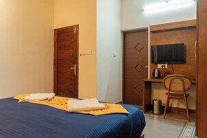 Quarto de hotel com cama, escrivaninha, cadeira, televisão e lixeira.