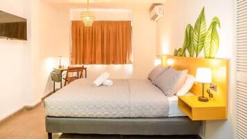 Quarto com cama, mesa de cabeceira com abajur, cadeira, TV fixada na parede e janela com cortinas.