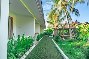 Um caminho leva a um edifício com varanda coberta, cercado por vegetação exuberante e uma palmeira.