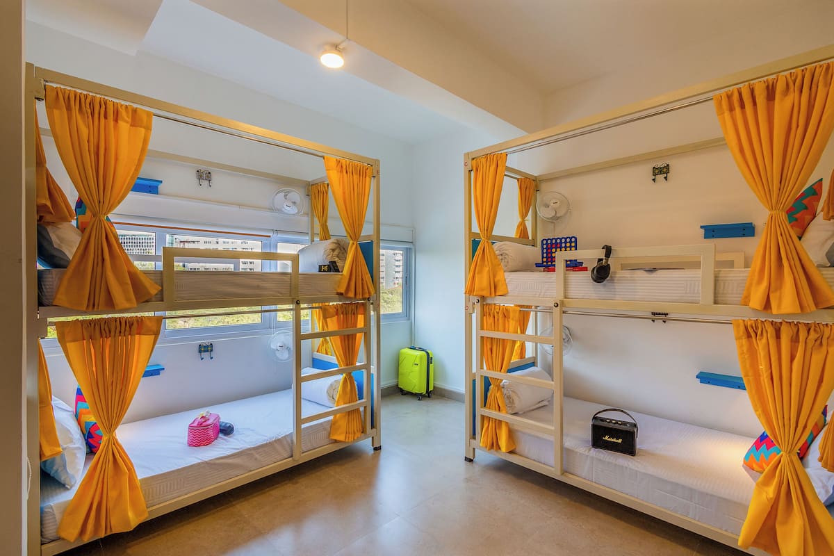 Um quarto de dormitório com beliches, cortinas, uma mala e um ventilador.