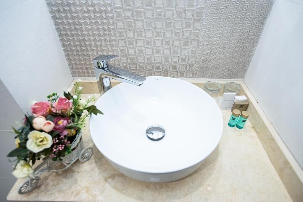 Uma casa de banho com uma pia branca arredondada, uma torneira cromada e um arranjo floral decorativo no lado esquerdo.
