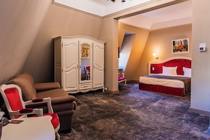 Quarto de hotel com cama vermelha, sofá marrom, guarda-roupa branco e poltrona vermelha.