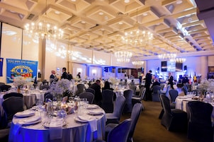 Uma sala de banquetes com mesas redondas preparadas para refeições, lustres e um palco com equipamentos musicais.