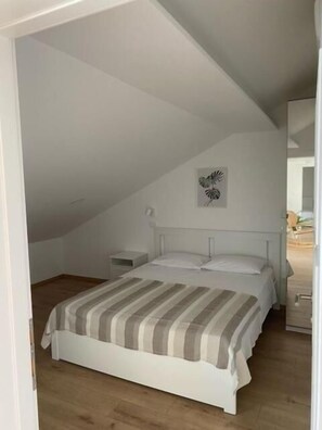 Quarto com teto inclinado, uma cama com colcha listrada, mesa de cabeceira e luminária fixada na parede.