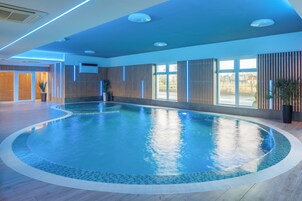 Uma piscina interior com borda em azulejo, soalho de madeira e amplas janelas que permitem entrada de luz natural.