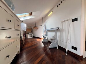 Quarto com piso de madeira, paredes brancas, cômoda branca, mesa pequena e radiador.