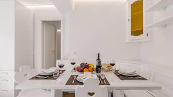 Uma mesa de jantar branca com quatro cadeiras, uma tigela de frutas, copos de vinho e uma garrafa de vinho.
