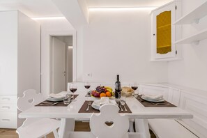 Uma mesa de jantar branca com quatro cadeiras, uma tigela de frutas, copos de vinho e uma garrafa de vinho.