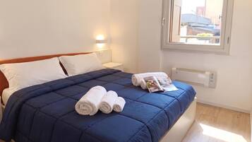 Cama bem arrumada com edredom azul, almofadas brancas e toalhas dobradas. Uma mesa de cabeceira com abajur e uma janela com vista para prédios.