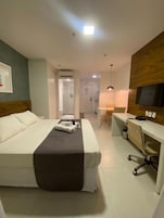 Quarto de hotel com cama, escrivaninha, cadeira, televisão e banheiro ao fundo.