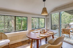 Uma sala de jantar com mesa de madeira posta para quatro pessoas, janelas amplas e vista para a floresta.