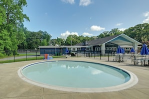 Piscina com área de estar coberta e guarda-sóis, cercada por uma grade e árvores.