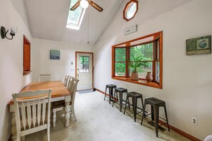 Uma sala de jantar com mesa de madeira e cadeiras brancas, ventilador de teto e janela com vista para o verde.