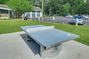 Mesa de tênis ao ar livre, com um edifício e carros estacionados ao fundo.