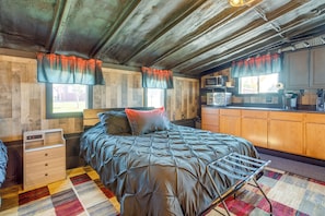 Quarto com cama, guarda-roupa de madeira, pequena escrivaninha e cozinha compacta.
