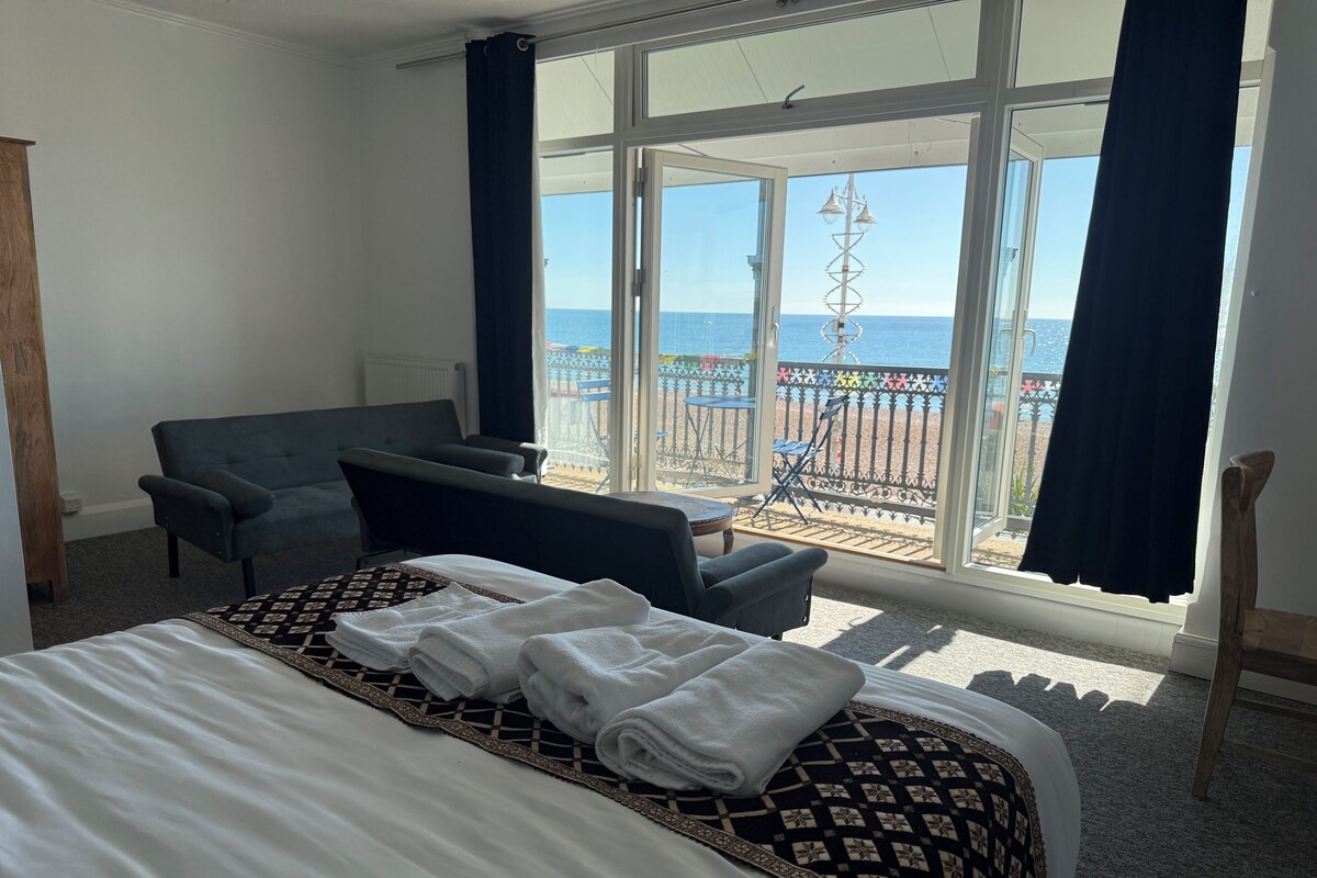 Quarto de hotel com cama, sofá, mesa e vista para o mar através de amplas janelas.