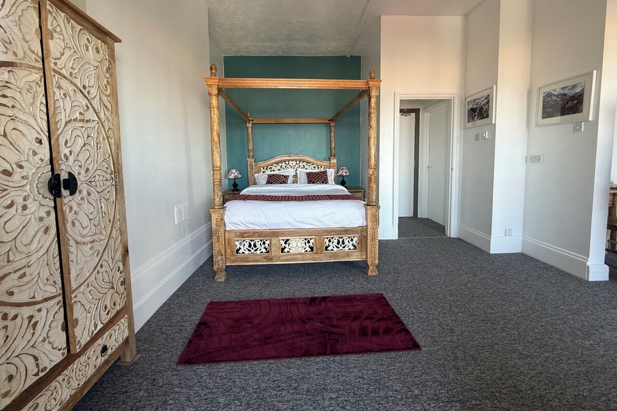 Uma cama de dossel com estrutura de madeira e detalhes entalhados, tapete vermelho escuro e parede cor verde-água.