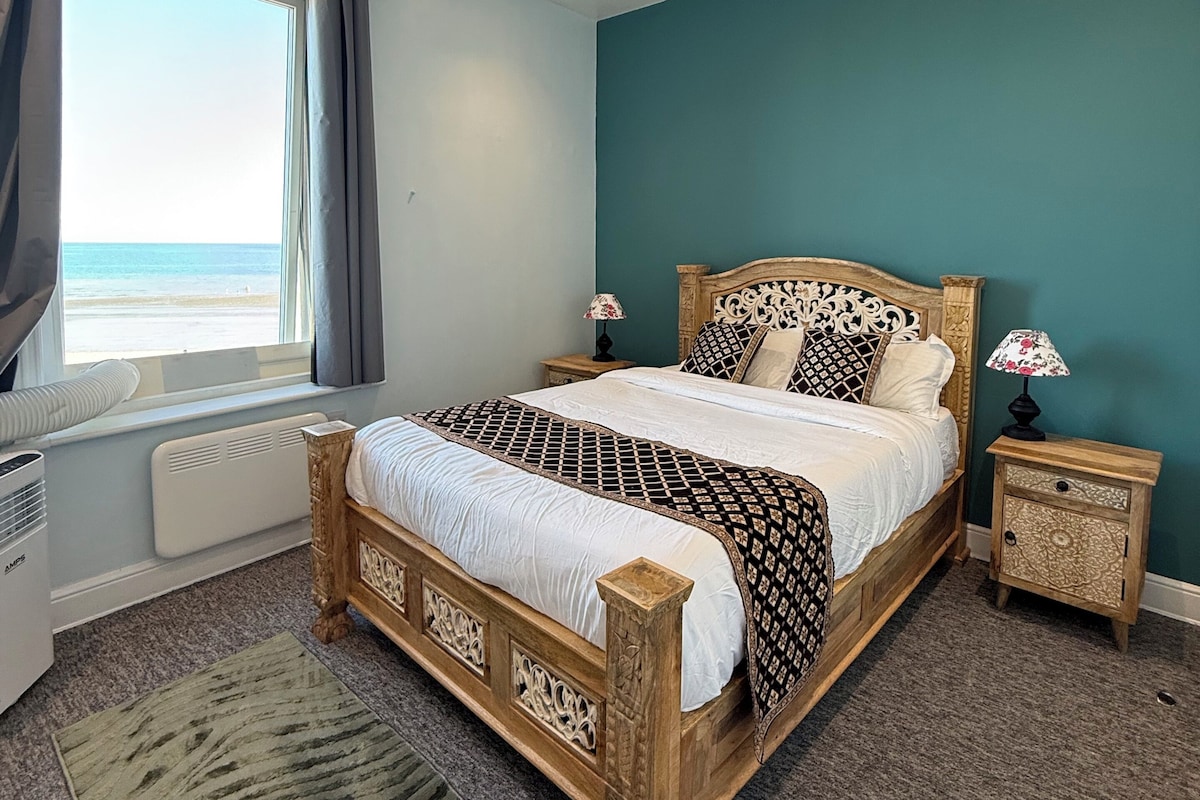 Quarto com cama, mesa de cabeceira, abajur, janela com cortinas e vista para a praia.
