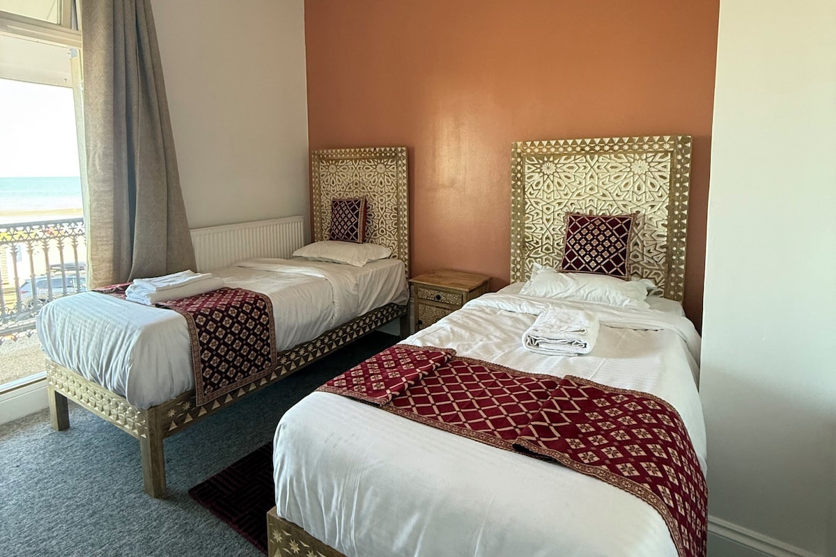 Quarto de hotel com duas camas, cada uma com cabeceira estampada, uma janela com cortinas e vista para o exterior.