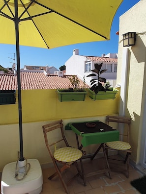 Uma pequena área de estar ao ar livre com uma mesa, duas cadeiras, um guarda-sol amarelo e plantas em vasos numa varanda.