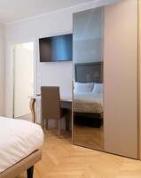 Quarto de hotel com cama, escrivaninha, cadeira, televisão e guarda-roupa.