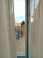 Vista pela janela, com porta, uma cadeira e um edifício com telhado de azulejos ao fundo.