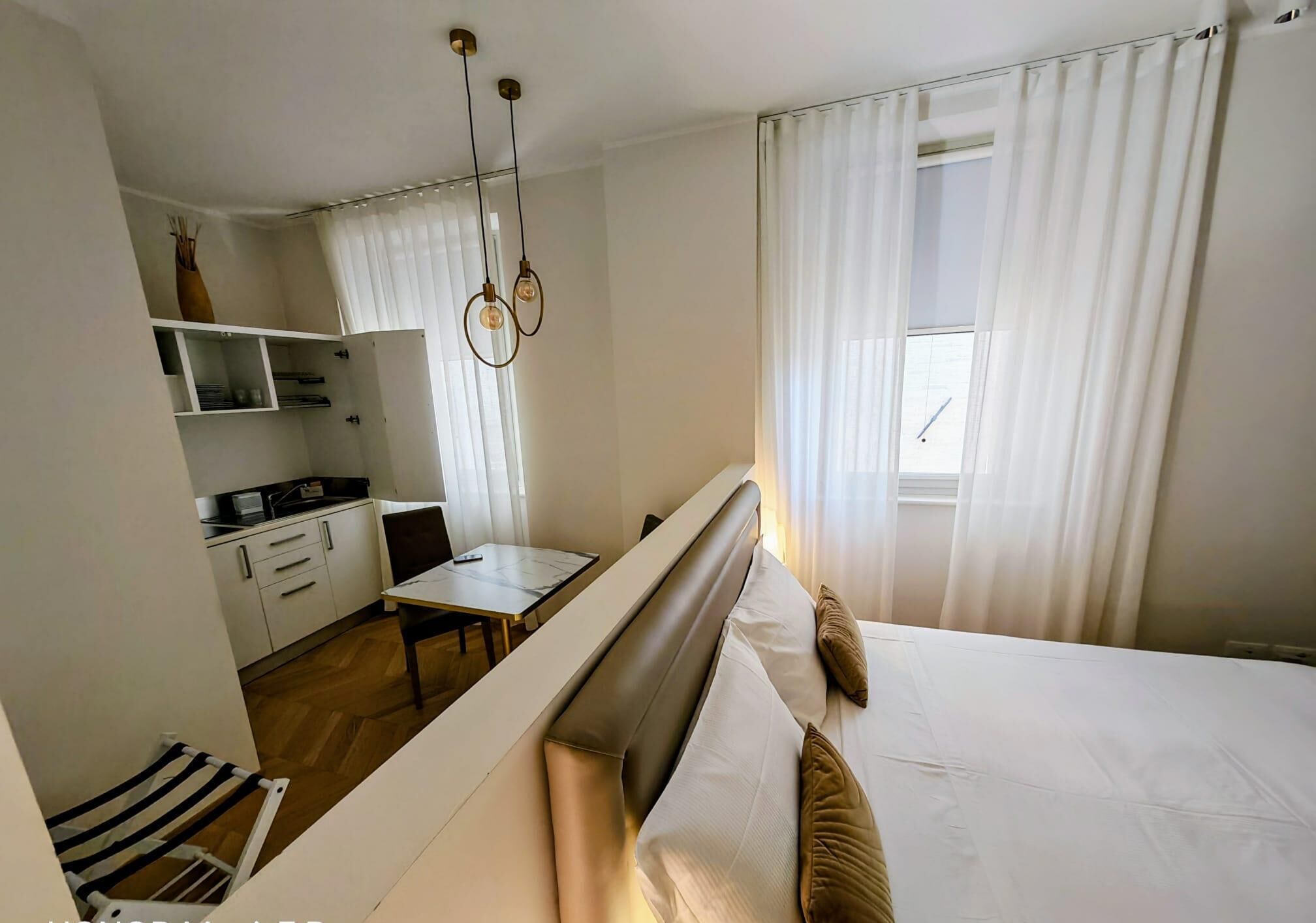 Quarto de hotel com cama, uma mesinha, cozinha compacta e janela com cortinas.