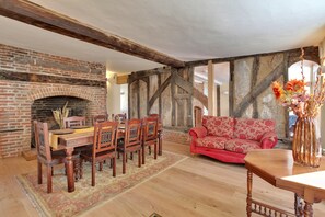 Um quarto com lareira de tijolos, mesa de jantar de madeira com cadeiras, um sofá floral vermelho e uma mesa de centro de madeira.