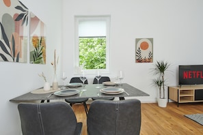 Uma sala de jantar com uma mesa posta para quatro pessoas, uma janela com vista para o verde e uma TV na parede exibindo o logotipo da Netflix.