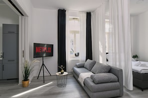 Uma sala de estar moderna com um sofá cinzento, uma mesa redonda pequena, uma TV preta sobre um tripé e vista para o exterior através de grandes janelas.