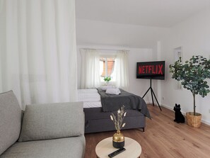 Quarto de hotel moderno com um sofá cinzento, uma cama com roupa de cama branca, uma televisão sobre um suporte e uma planta em vaso.