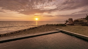 Área da piscina com vista para o pôr do sol sobre o oceano, uma espreguiçadeira e um pequeno jardim.