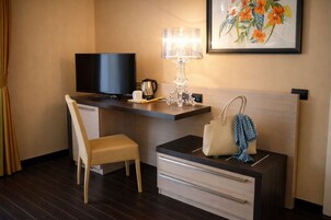Quarto de hotel com televisão de tela plana, escrivaninha com luminária, cadeira, mesa pequena, bolsa de mão e um quadro na parede.