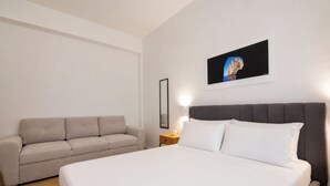 Quarto de hotel com uma cama, um sofá cinzento, uma mesinha de cabeceira com abajur e um quadro na parede.