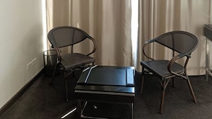 Quarto pequeno com duas cadeiras, uma mesa, cortinas bege e uma luminária no teto.