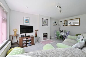 Uma sala de estar com televisão de tela plana, estante de livros, lareira, área de jantar com mesa e cadeiras, e um espelho grande na parede.