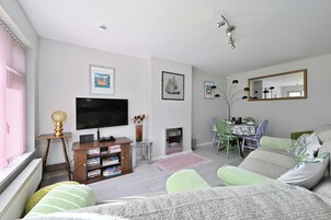 Uma sala de estar com televisão de tela plana, estante de livros, lareira, área de jantar com mesa e cadeiras, e um espelho grande na parede.