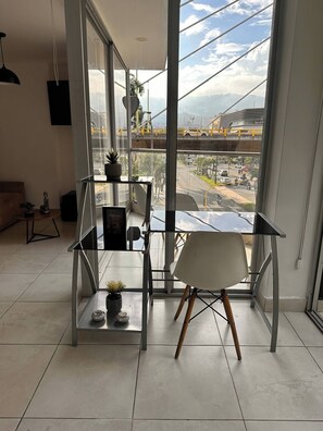 Quarto moderno com mesa e cadeira de vidro, um vaso de plantas e vista para a cidade através de amplas janelas.