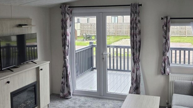 Quarto com televisão sobre um móvel, lareira, porta de vidro deslizante para uma varanda e cortinas com padrão de folhas.