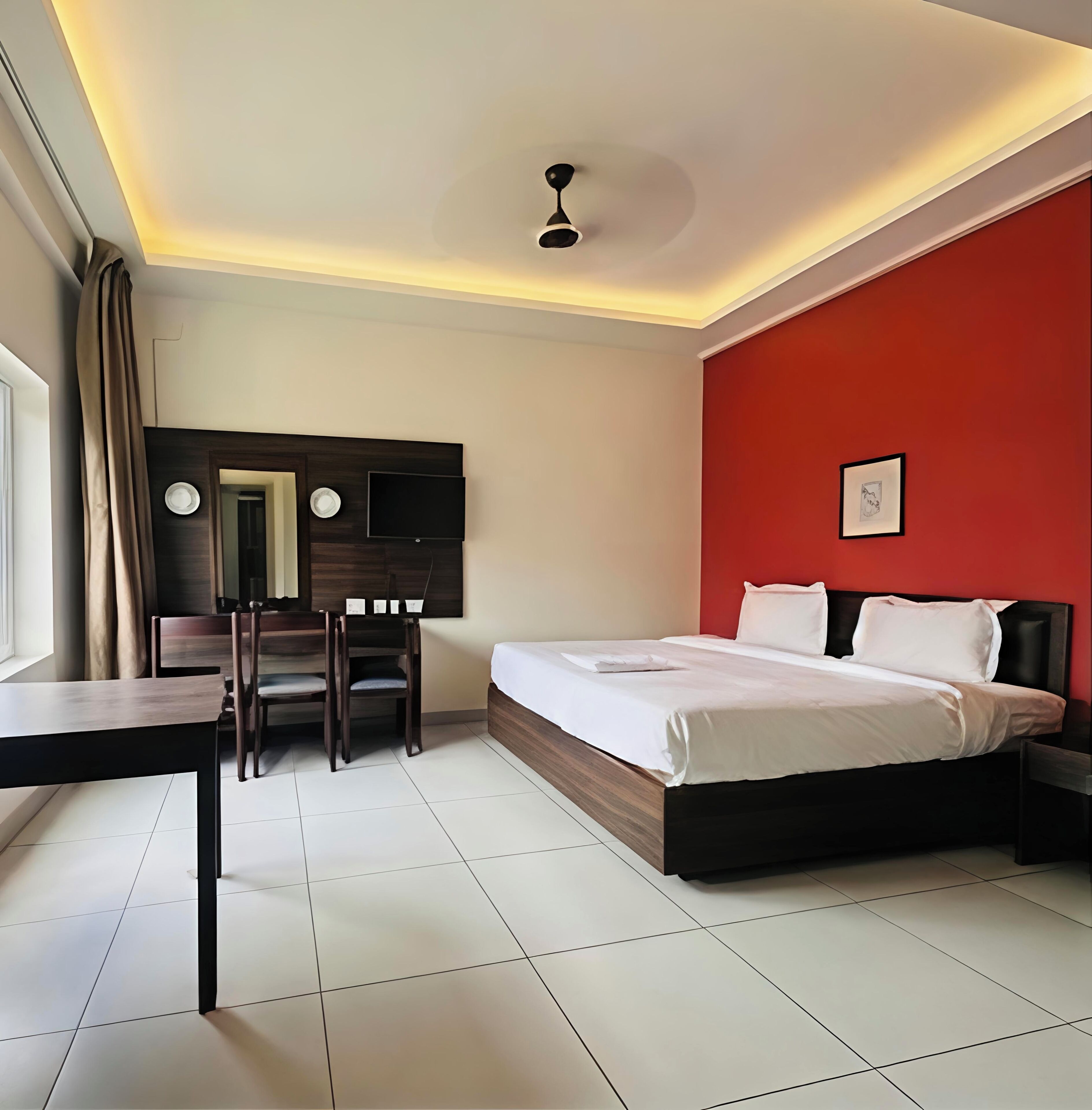Quarto de hotel com cama, escrivaninha, cadeira, televisão e uma parede de destaque vermelha.