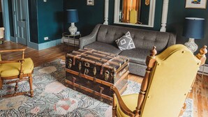 A room with a sofa, a yellow armchair, a vintage trunk, and a chandelier.