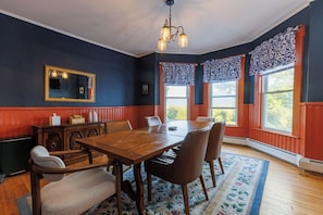 Uma sala de jantar com mesa e cadeiras de madeira, um lustre e janelas com cortinas estampadas.