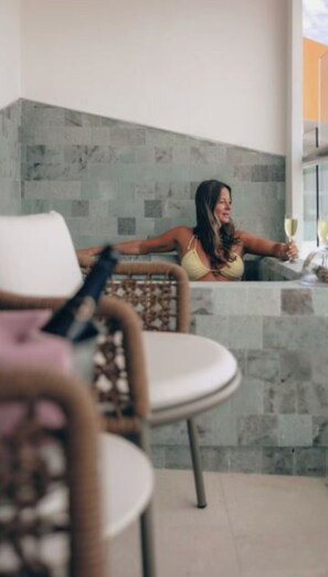 Mulher relaxando em uma banheira com uma toalha, rodeada por cadeiras e uma parede de pedra.