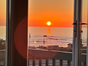 Vista do pôr do sol por uma janela com corrimão, uma bandeira e um corpo de água.