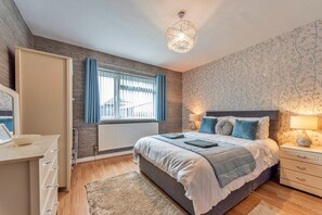 Quarto com cama, mesas de cabeceira, janela com cortinas, radiador e um lustre.